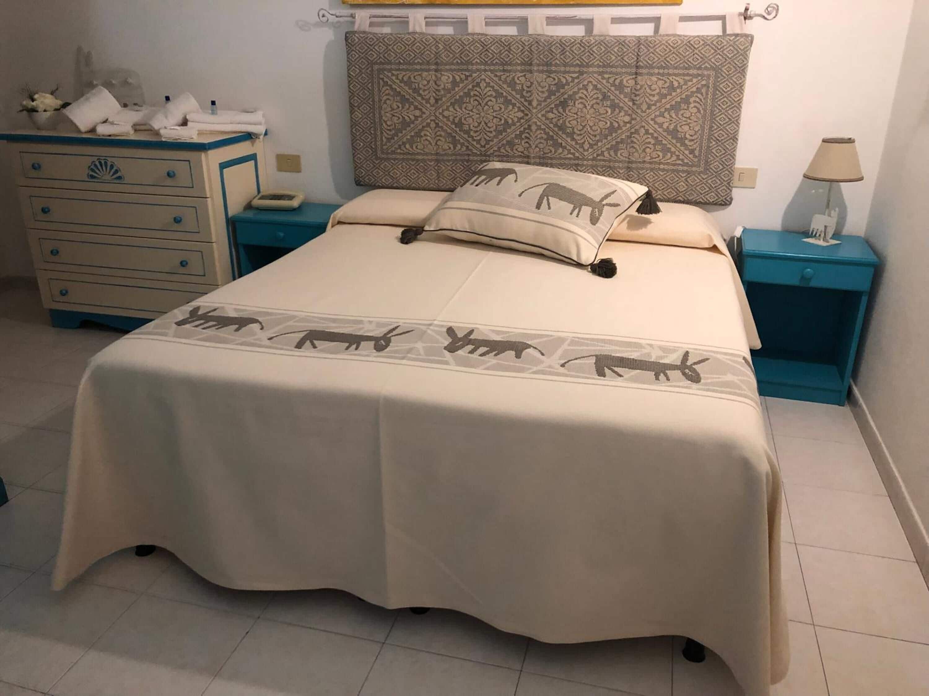Uma cama bem arrumada com colcha estampada, uma mesinha de cabeceira com abajur, uma cômoda com gavetas e um enfeite de parede estampado.