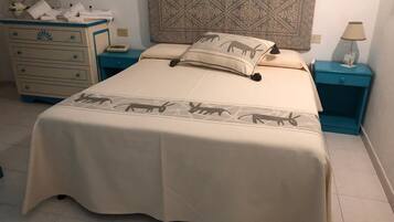 Uma cama bem arrumada com colcha estampada, uma mesinha de cabeceira com abajur, uma cômoda com gavetas e um enfeite de parede estampado.