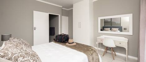 Um quarto com cama, escrivaninha, guarda-roupa e espelho.