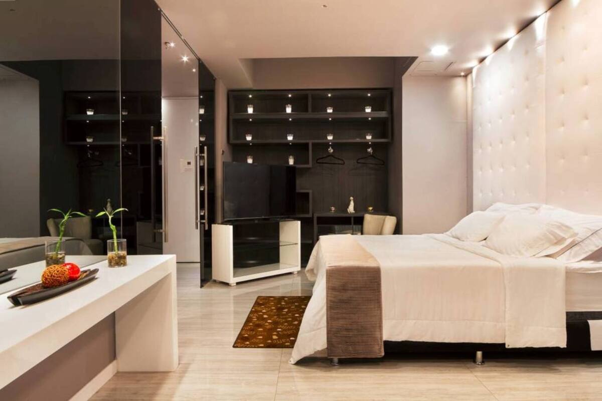 Quarto moderno com cama grande, mesa de cabeceira, espelho, guarda-roupa com espaço para pendurar roupas e um pequeno espaço de estar.