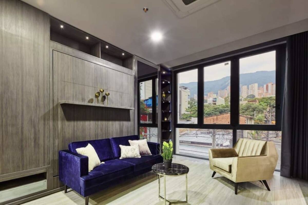 Uma sala de estar moderna com um sofá azul, uma poltrona bege e uma grande janela com vista para a cidade.