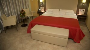 Quarto de hotel com uma cama grande, uma mesa de cabeceira com abajur, uma cadeira e uma pequena mesa com um vaso de flores.