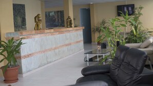 Uma área de recepção com balcão de mármore, duas estátuas douradas, uma televisão e vários vasos de plantas.