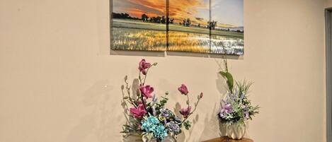 Um quarto com uma mesa de madeira, dois vasos com flores e uma pintura de paisagem ao pôr do sol na parede.
