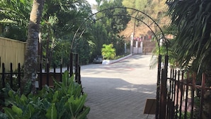 Uma trilha pavimentada leva a um portão com um arco de metal, tudo cercado por vegetação exuberante e palmeiras.
