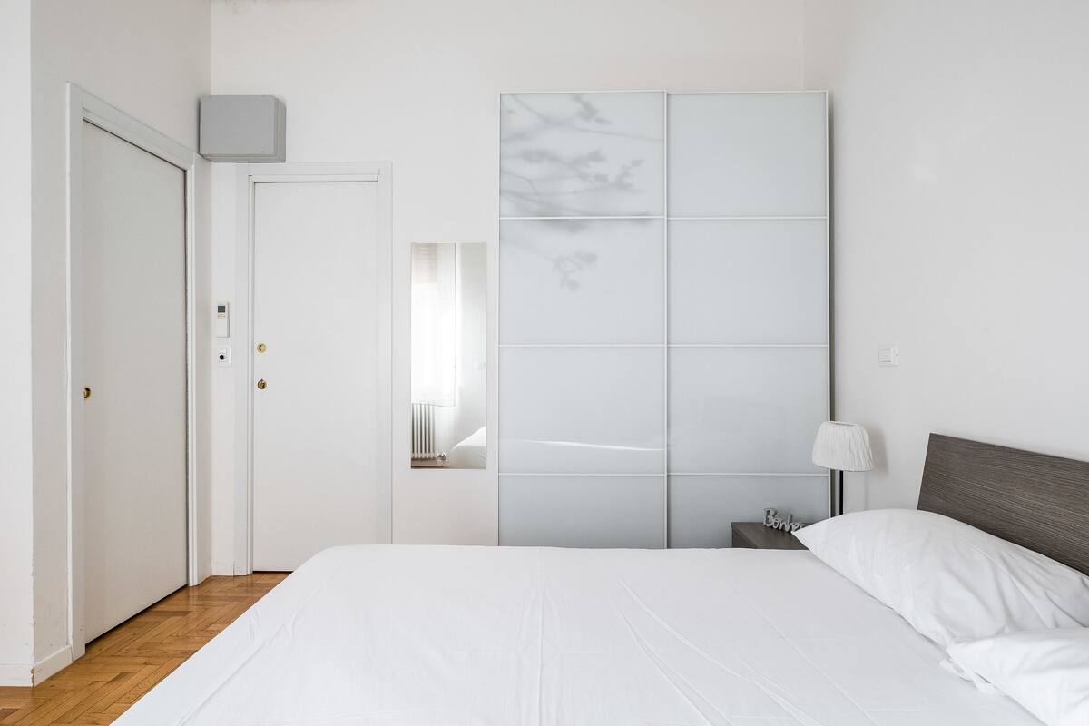 Quarto com uma cama, mesa de cabeceira, luminária, guarda-roupa e uma porta.