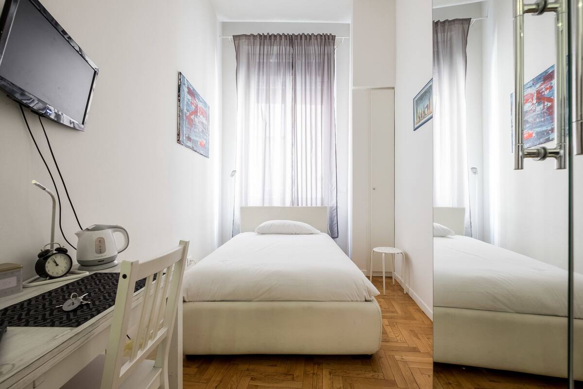 Quarto de hotel com duas camas, uma escrivaninha com chaleira, relógio e uma mesinha.