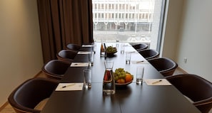 Sala de conferências com uma mesa comprida, cadeiras, copos de água, uma cesta de frutas e uma janela com vista para um edifício.