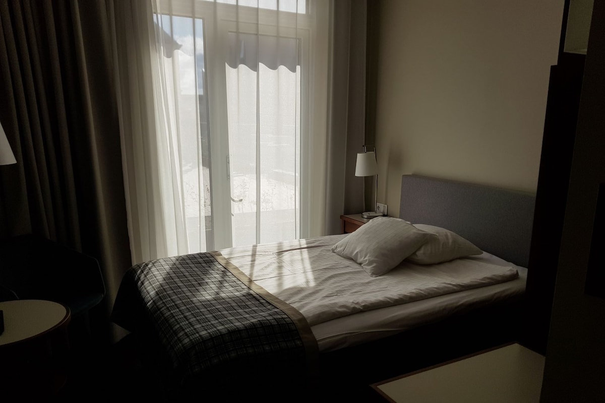 Quarto de hotel com cama, mesa de cabeceira com luminária e janela com cortinas.