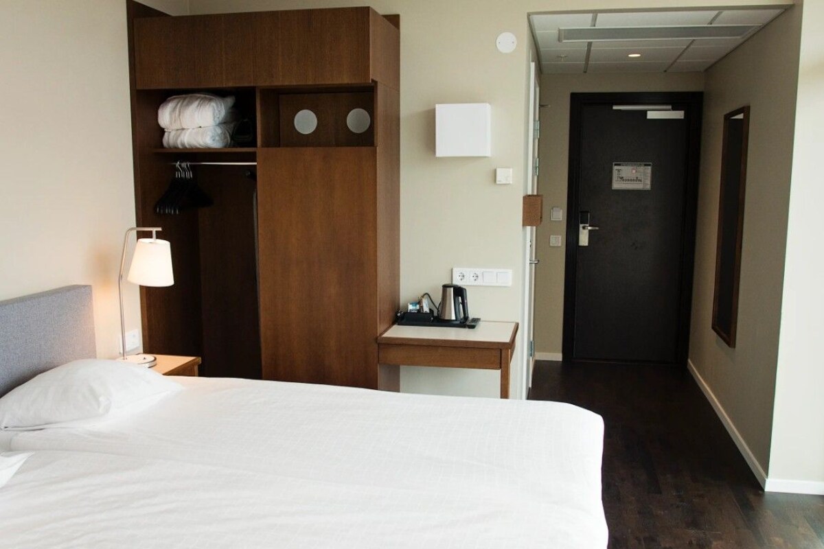 Quarto de hotel com cama, escrivaninha com cafeteira, armário e porta que dá para outro espaço.