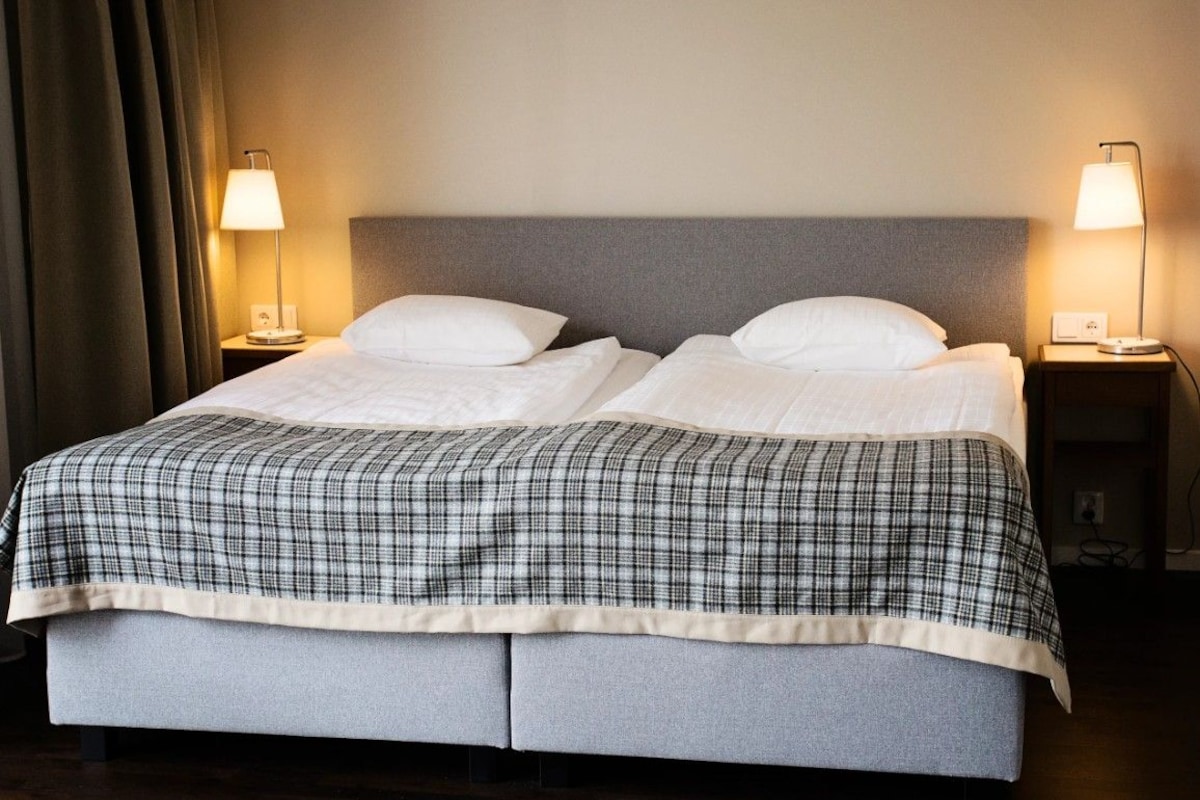 Uma cama de casal com cabeceira cinzenta, duas luminárias de cabeceira e um edredom xadrez.