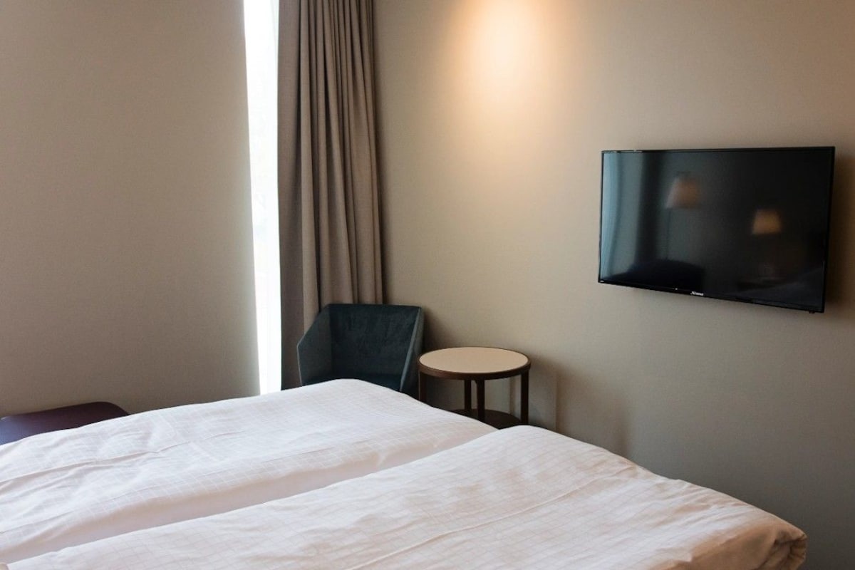 Quarto de hotel com cama, uma cadeira, mesa redonda pequena e televisão de tela plana fixada na parede.