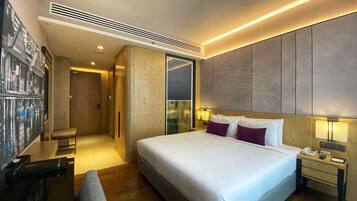 Quarto de hotel moderno com uma cama grande, uma escrivaninha, uma cadeira e um banheiro visível através de uma porta de vidro.