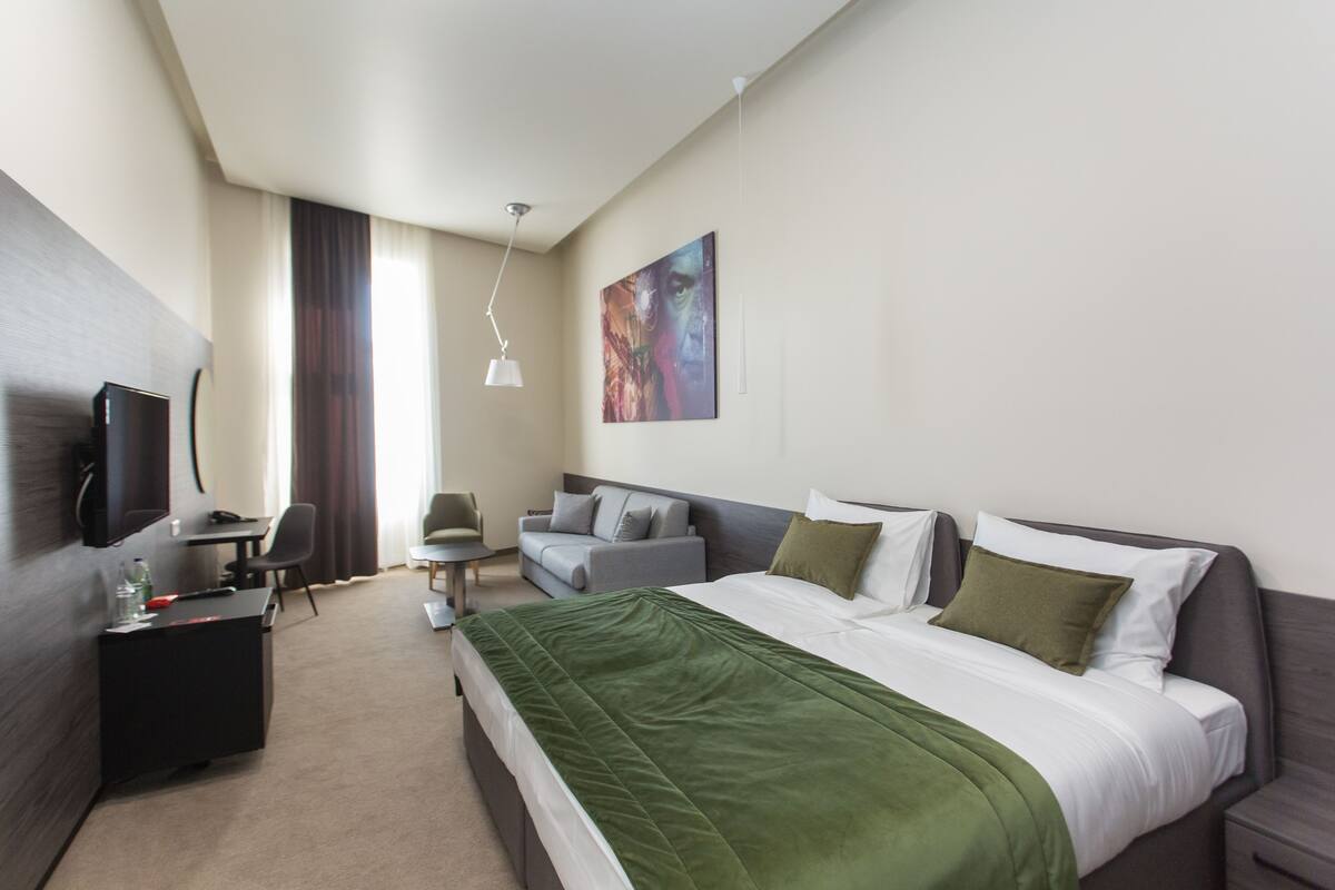 Quarto de hotel com cama, escrivaninha, cadeira, televisão e um quadro na parede.