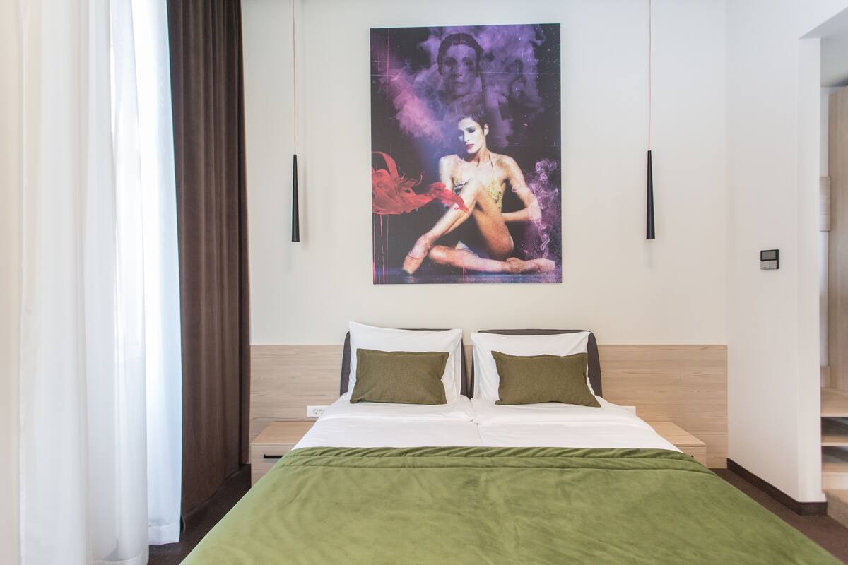 Quarto de hotel moderno com uma cama grande, duas almofadas, um cobertor verde e uma obra de arte de grandes dimensões na parede.
