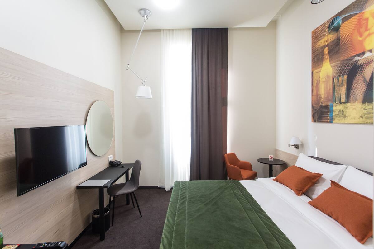 Quarto de hotel com televisão de tela plana, escrivaninha com cadeira, cama com roupa de cama verde e laranja, e um quadro na parede.