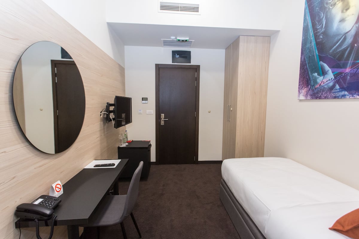 Quarto de hotel com cama, escrivaninha com telefone, espelho, porta e projetor fixado na parede.