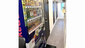 Máquina de vendas com diversas bebidas e lanches, localizada em um corredor com portas e piso coberto de carpete.