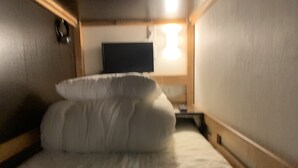 Um quarto pequeno e com pouca luz, com uma cama, uma televisão de tela plana na parede e cabeceira de madeira.
