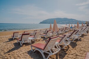 Uma praia com fileiras de espreguiçadeiras e guarda-sóis, um mar calmo e uma colina ao fundo.