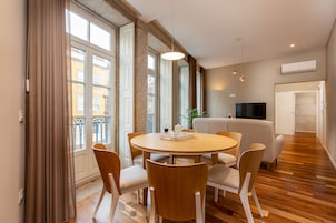 Uma área de jantar moderna com uma mesa redonda e seis cadeiras, janelas amplas e uma TV ao fundo.