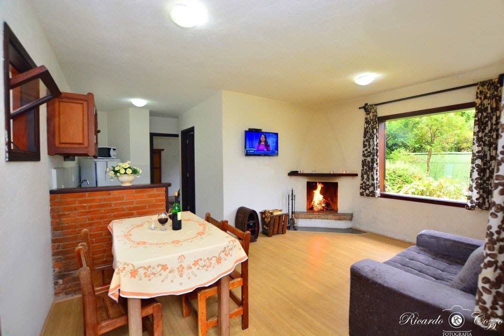 Uma sala de estar acolhedora com lareira, um espaço de jantar com mesa posta para dois e uma televisão fixada na parede.