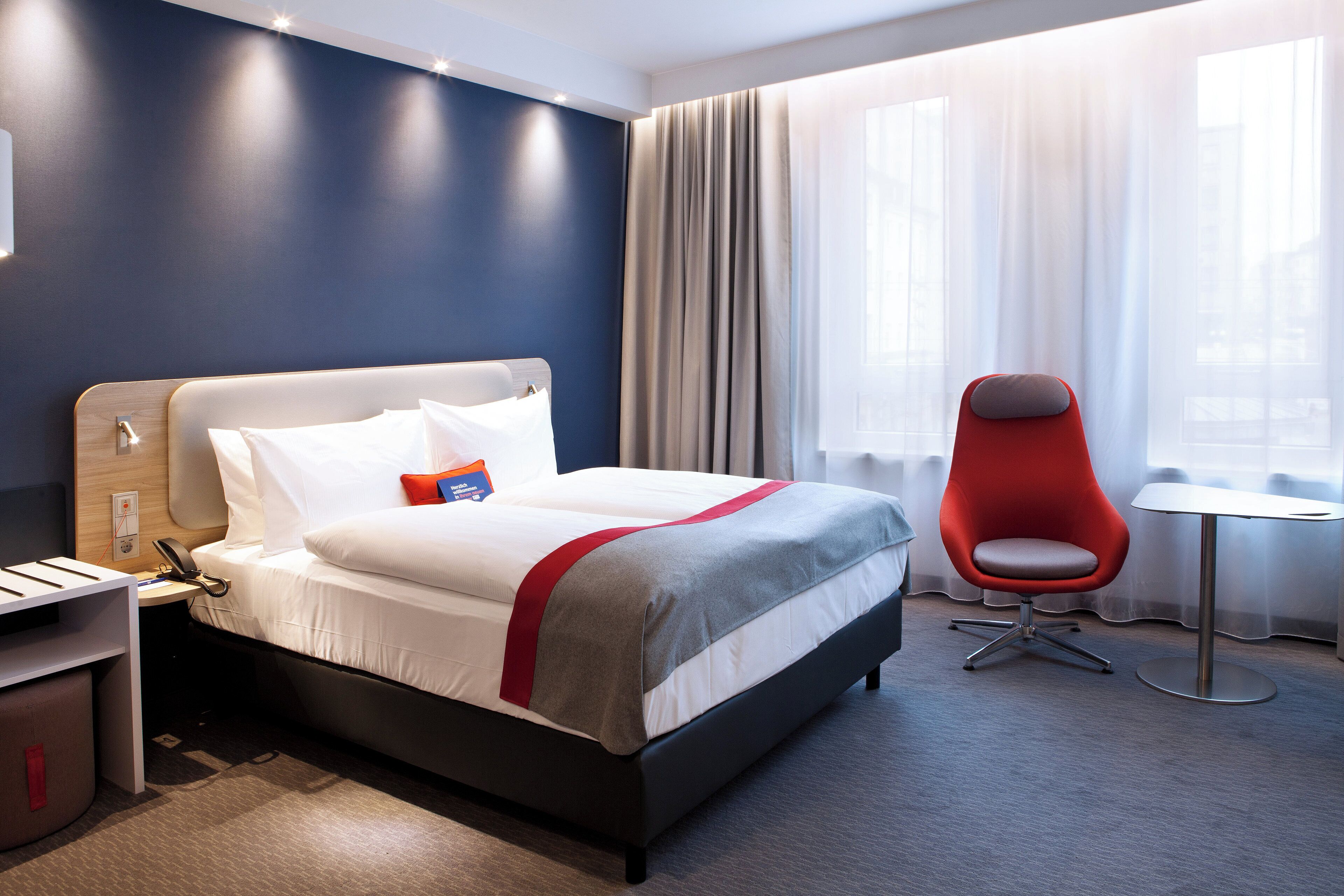 Quarto de hotel com cama, escrivaninha, cadeira e janela com cortinas.