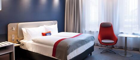 Quarto de hotel com cama, escrivaninha, cadeira e janela com cortinas.