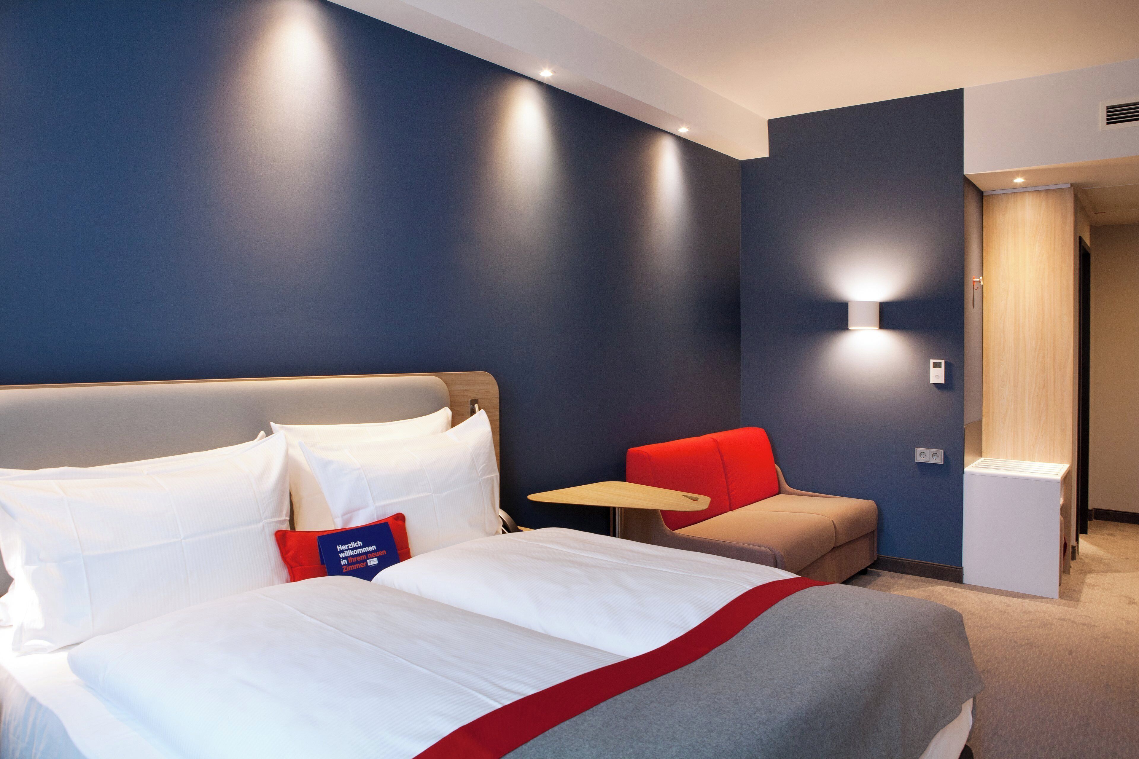 Quarto de hotel com cama, uma mesinha, uma cadeira vermelha e uma luminária fixada na parede.