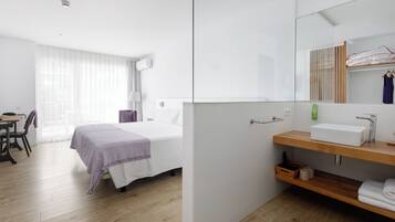 Quarto de hotel com cama, mesa de cabeceira com espelho, prateleira de madeira com produtos de higiene pessoal e janela com cortinas finas.