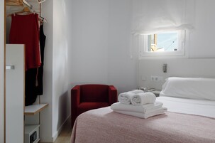 Quarto de hotel com uma cama, uma poltrona vermelha, um guarda-roupa e uma janela com vista para um edifício.