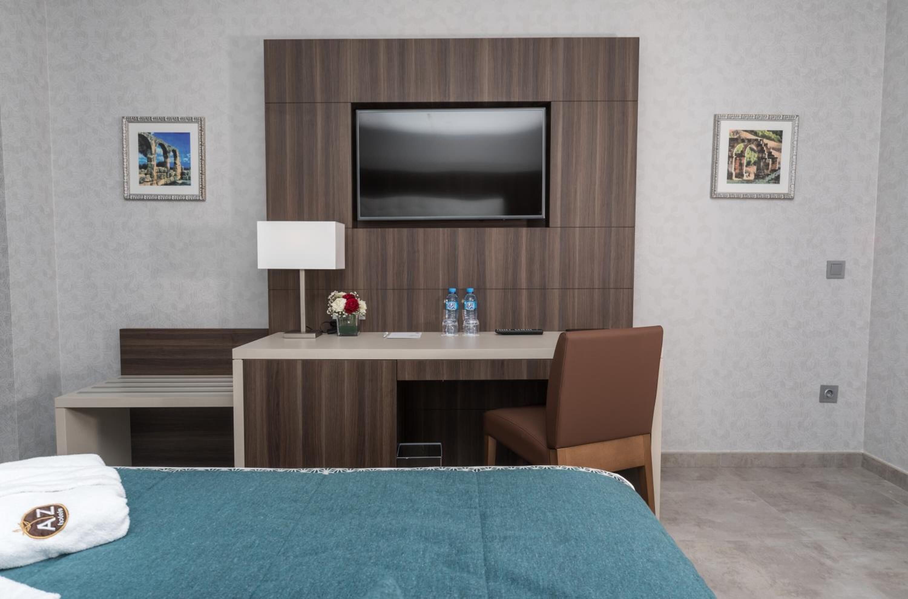 Quarto de hotel com uma cama, uma secretária com uma TV, uma cadeira, um abajur e duas imagens emolduradas nas paredes.