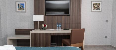 Quarto de hotel com uma cama, uma secretária com uma TV, uma cadeira, um abajur e duas imagens emolduradas nas paredes.