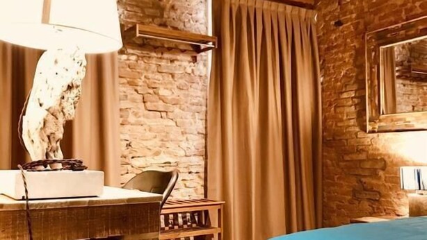 Um quarto com teto de madeira, parede de pedra, uma cama com colcha azul, uma escrivaninha com abajur e uma cadeira.