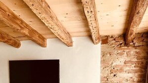 Quarto com vigas de madeira aparentes, parede de tijolos, televisão de tela plana, escrivaninha com cadeira, luminária e um pequeno frigorífico.