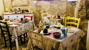 Uma área de jantar rústica com parede de pedra como cenário, cadeiras de madeira, mesa posta com diversos pratos e utensílios, e uma geladeira antiga.