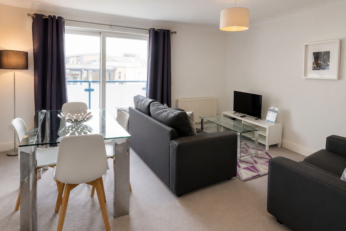 Sala de estar moderna com mesa de jantar de vidro, dois sofás, televisão e vista para a cidade.
