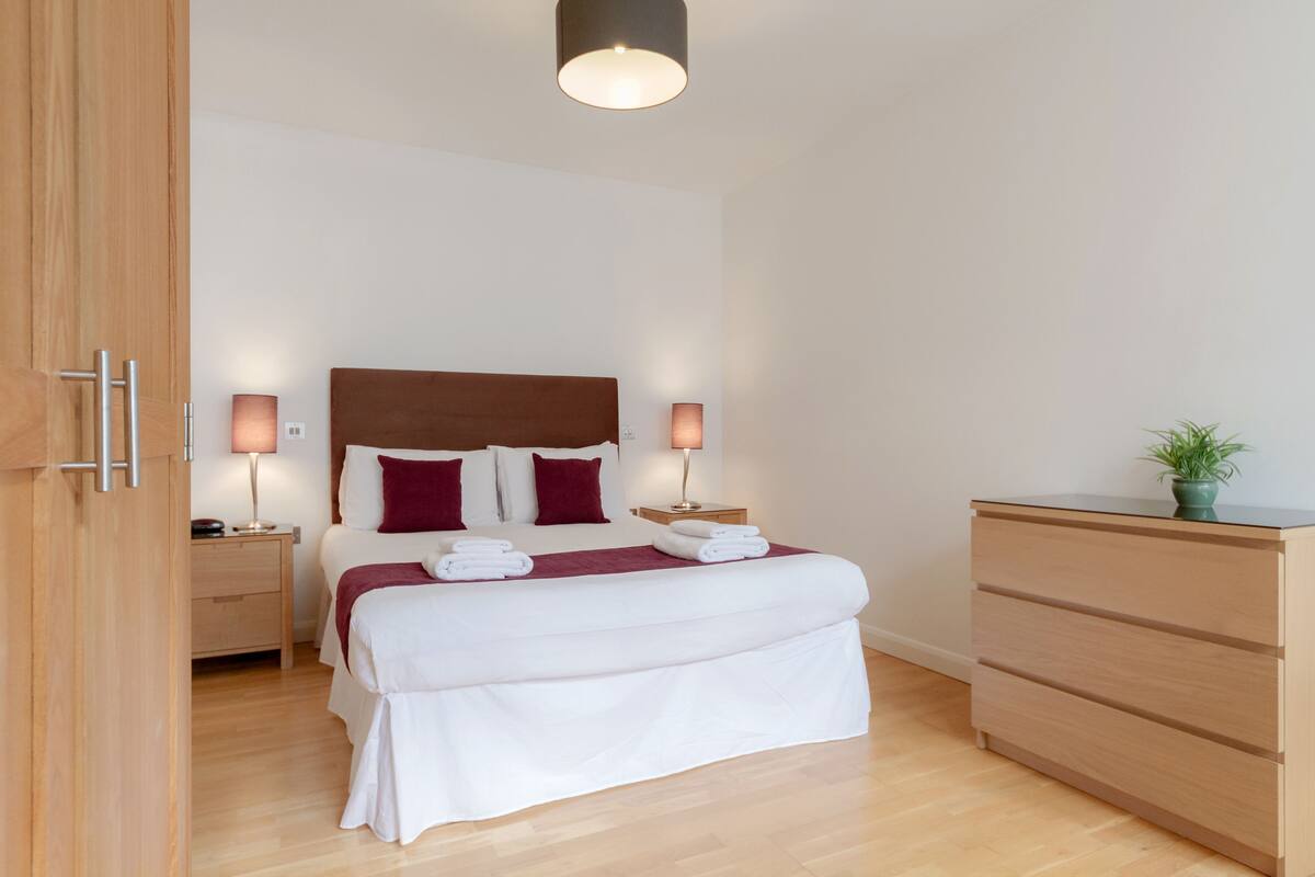 Quarto de hotel bem arrumado com cama, mesas de cabeceira, penteadeira e luminária.