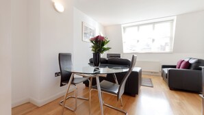 Uma sala de estar moderna com uma mesa de jantar de vidro, cadeiras pretas, um sofá cinzento e uma janela com persianas.