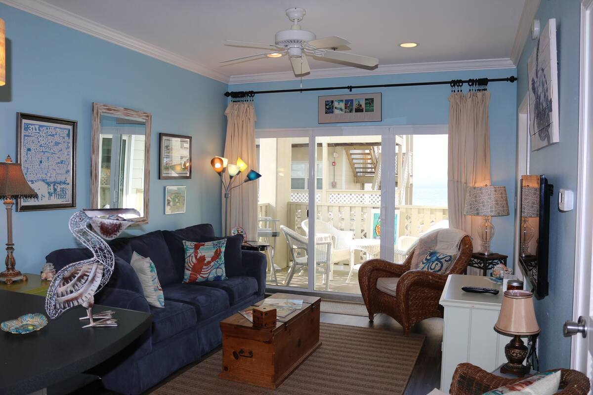 Sala de estar com sofá azul, um baú de madeira e uma cadeira de vime. Há um ventilador de teto, uma mesa com abajur e vista para o exterior.