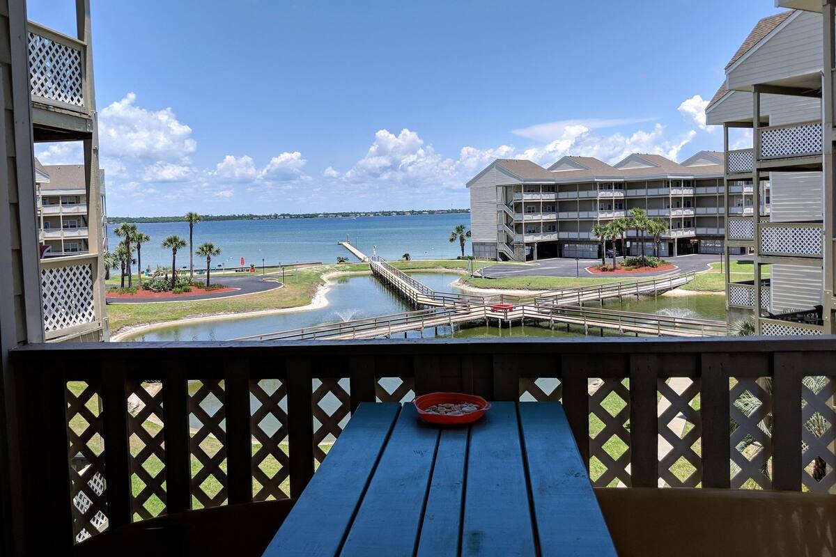 Vista de uma varanda com vista para uma marina, incluindo um cais, palmeiras e um edifício com várias varandas.