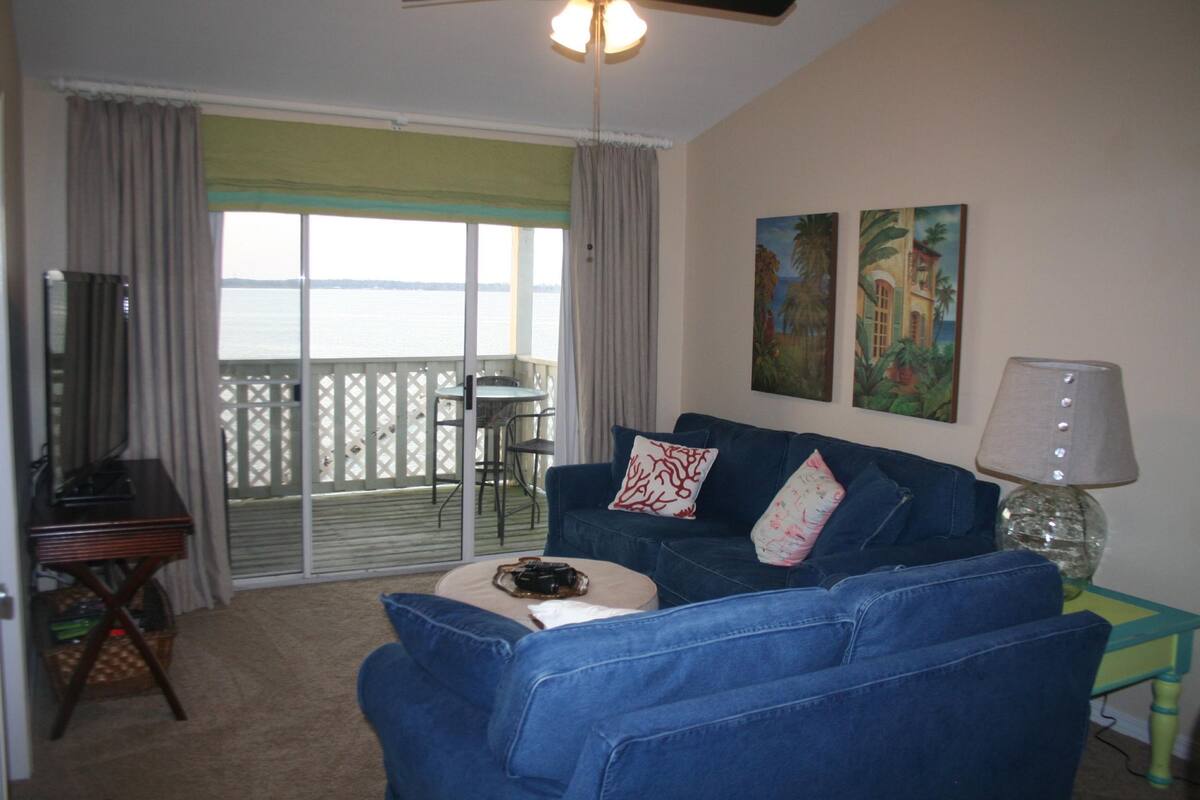 Uma sala de estar com um sofá azul, uma mesinha redonda com um objeto decorativo, e vista para o mar através de uma porta de vidro.