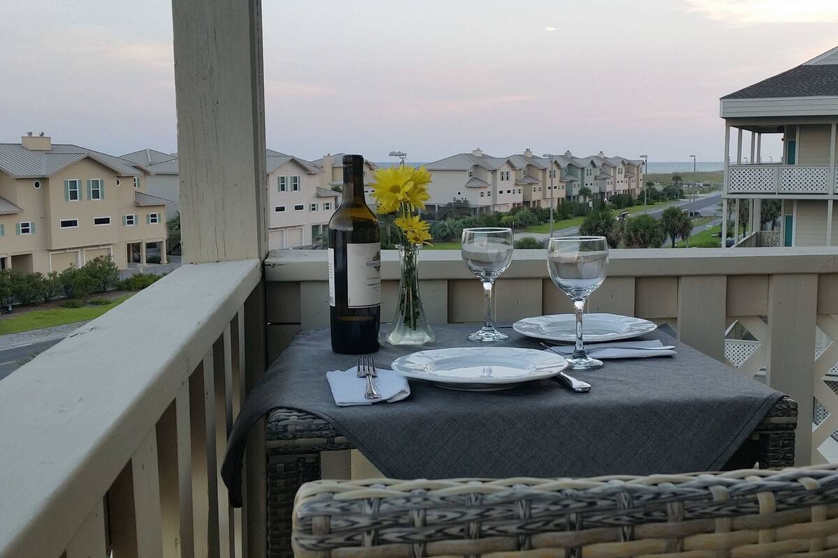 Mesa de varanda posta com taças de vinho, garrafa e um vaso com flores amarelas.