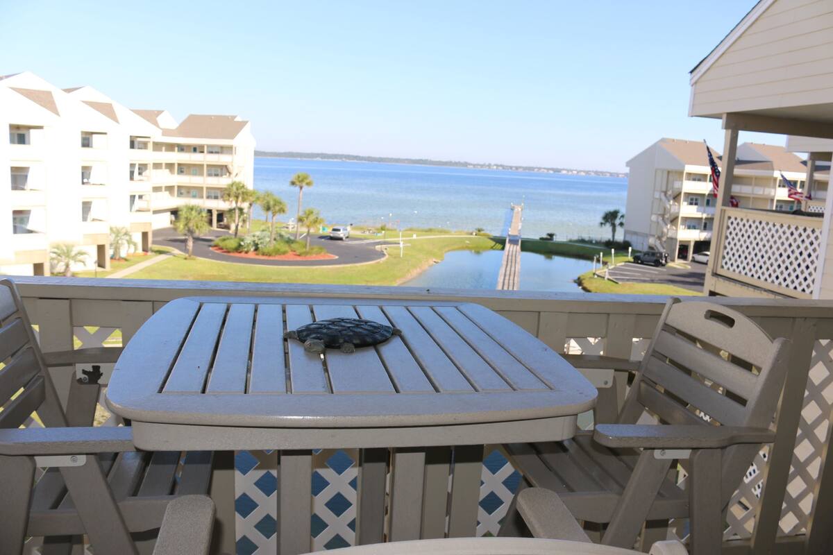 Varanda com mesa e cadeiras, vista para um corpo de água e edifícios.
