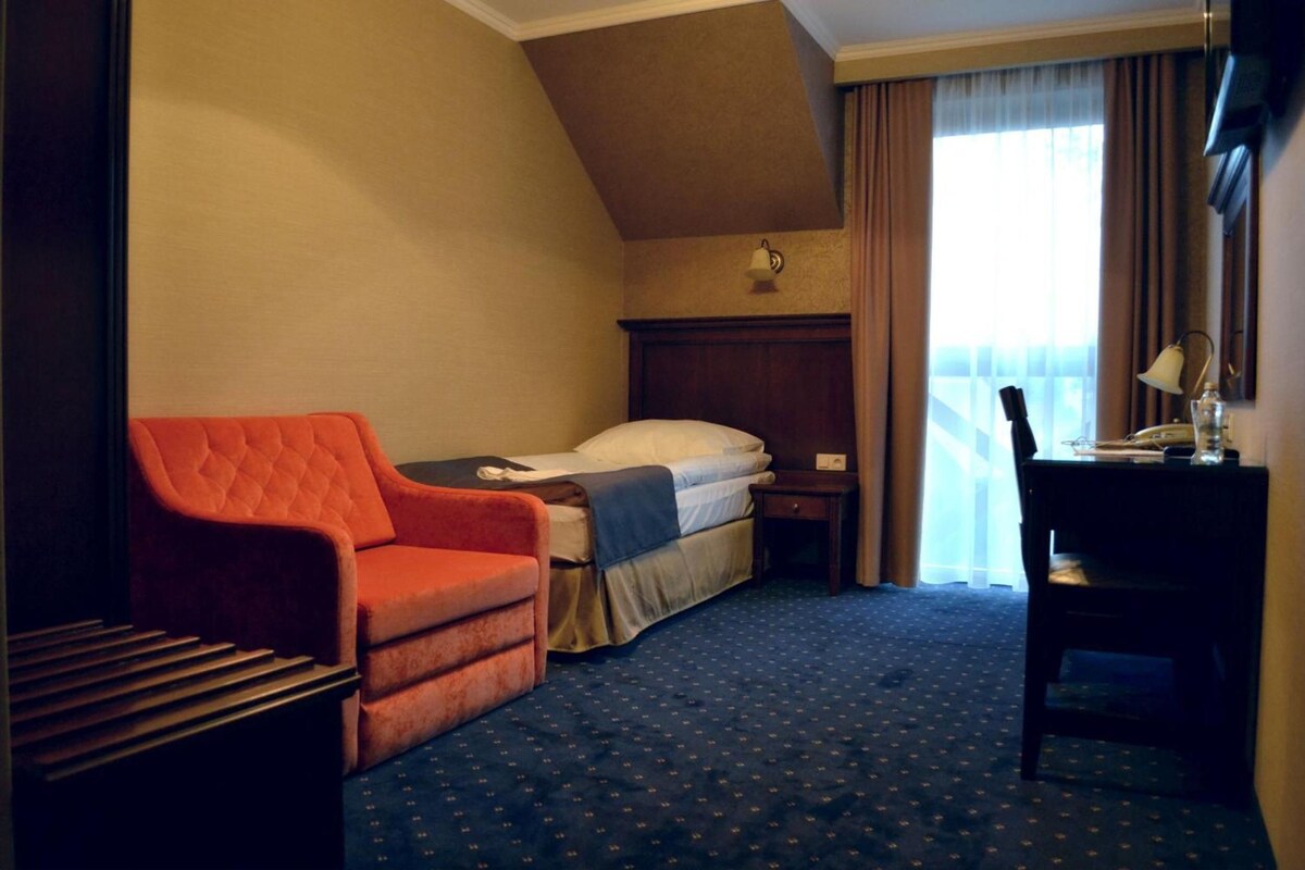 Quarto de hotel com uma cama de solteiro, uma poltrona vermelha, uma escrivaninha com luminária e uma janela com cortinas.