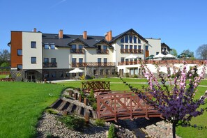 Um hotel de vários andares com fachada de pedra, área de estar externa e jardim com uma ponte de madeira.