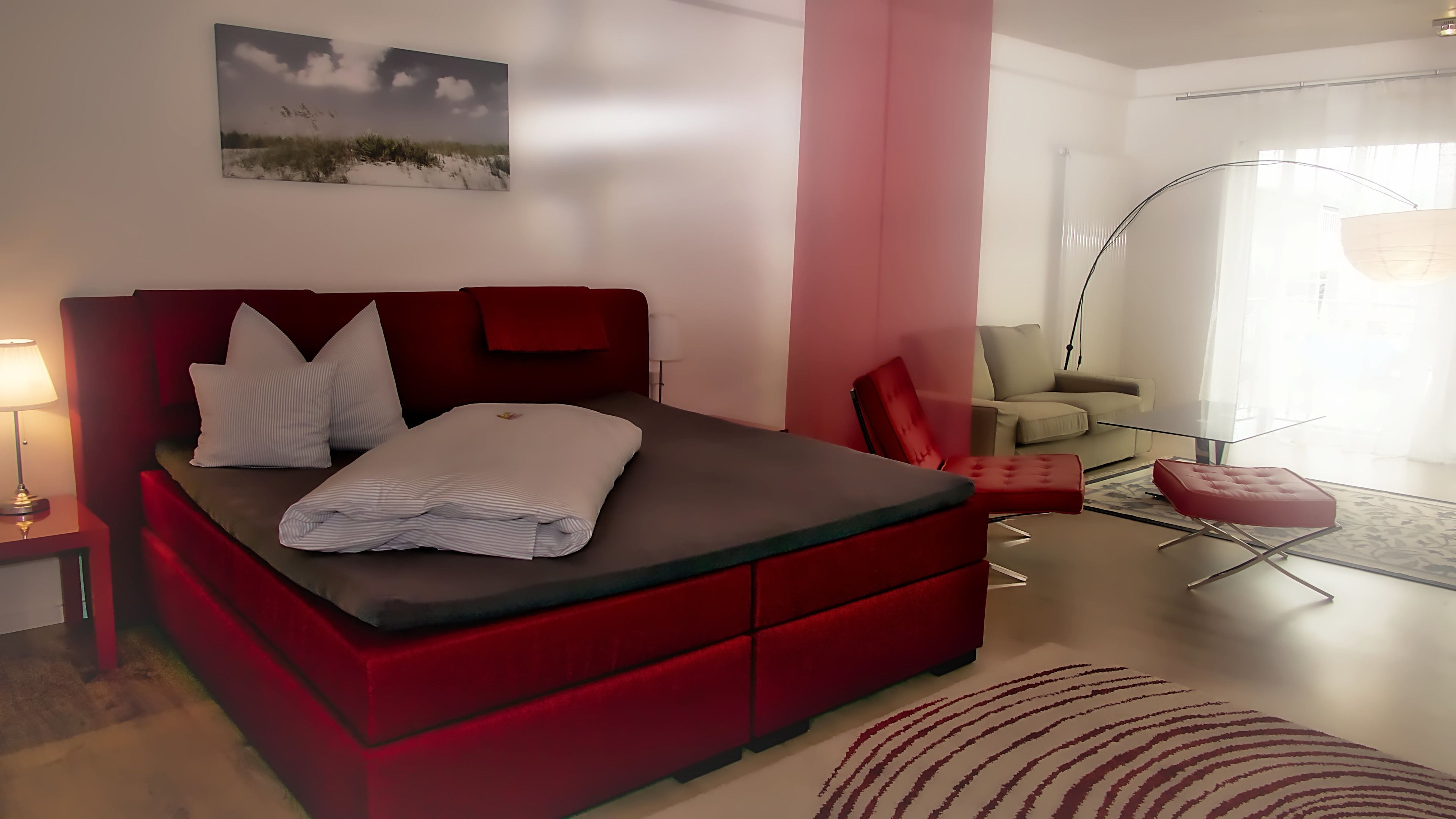 Um quarto moderno com uma cama vermelha, uma mesinha de cabeceira com abajur, uma cadeira e uma mesa de vidro.