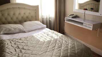 Uma cama com cabeceira acolchoada, mesa de cabeceira com telefone e um vaso, e um espelho em cima.