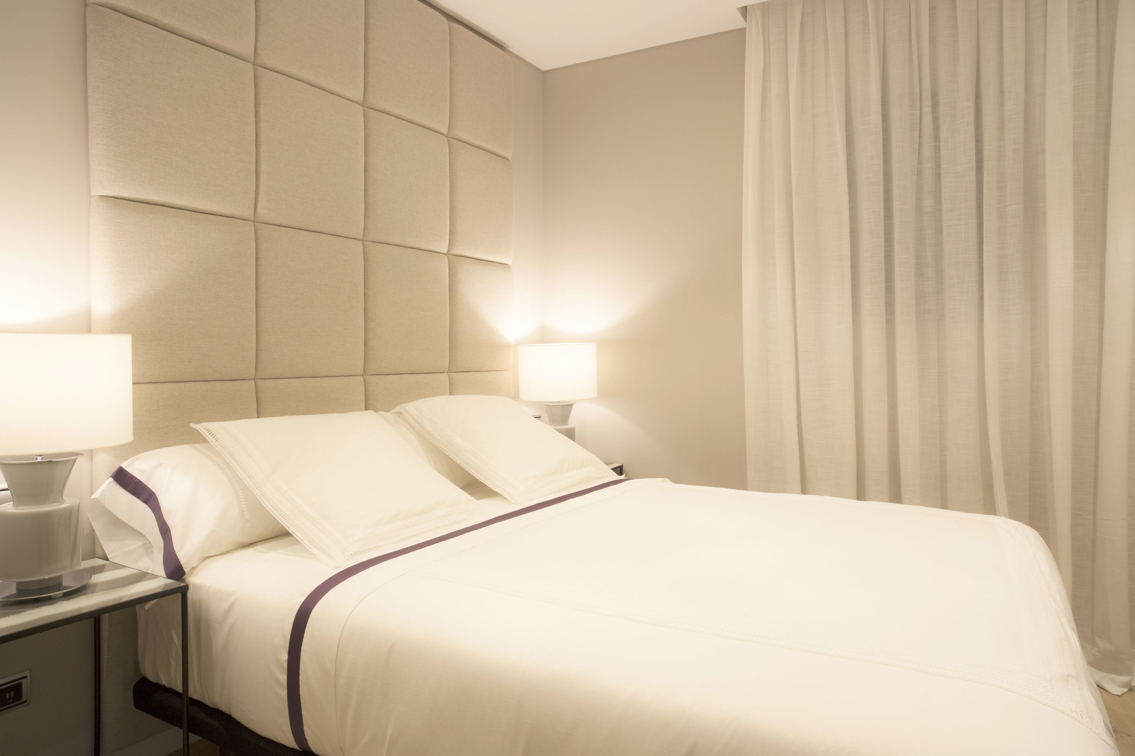 Quarto de hotel com uma cama grande, duas luminárias de cabeceira, mesa de apoio e janela com cortinas.