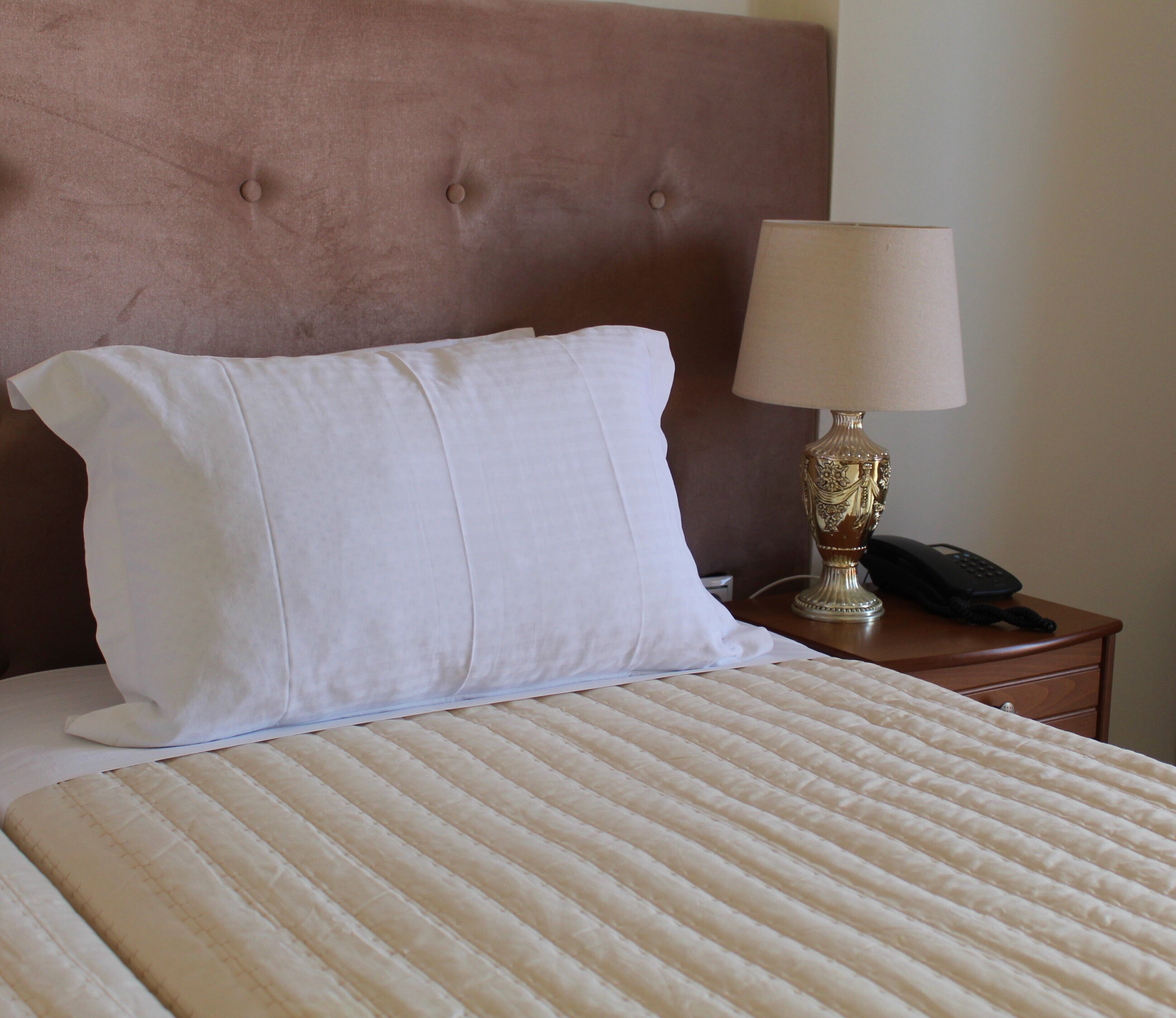 Cama bem arrumada com travesseiro branco e manta acolchoada bege. Um criado-mudo de madeira com abajur e telefone fica ao lado da cama.